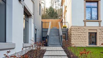Um edifício moderno com uma escadaria que leva a uma porta, rodeado por um jardim com plantas e um caminho pavimentado.
