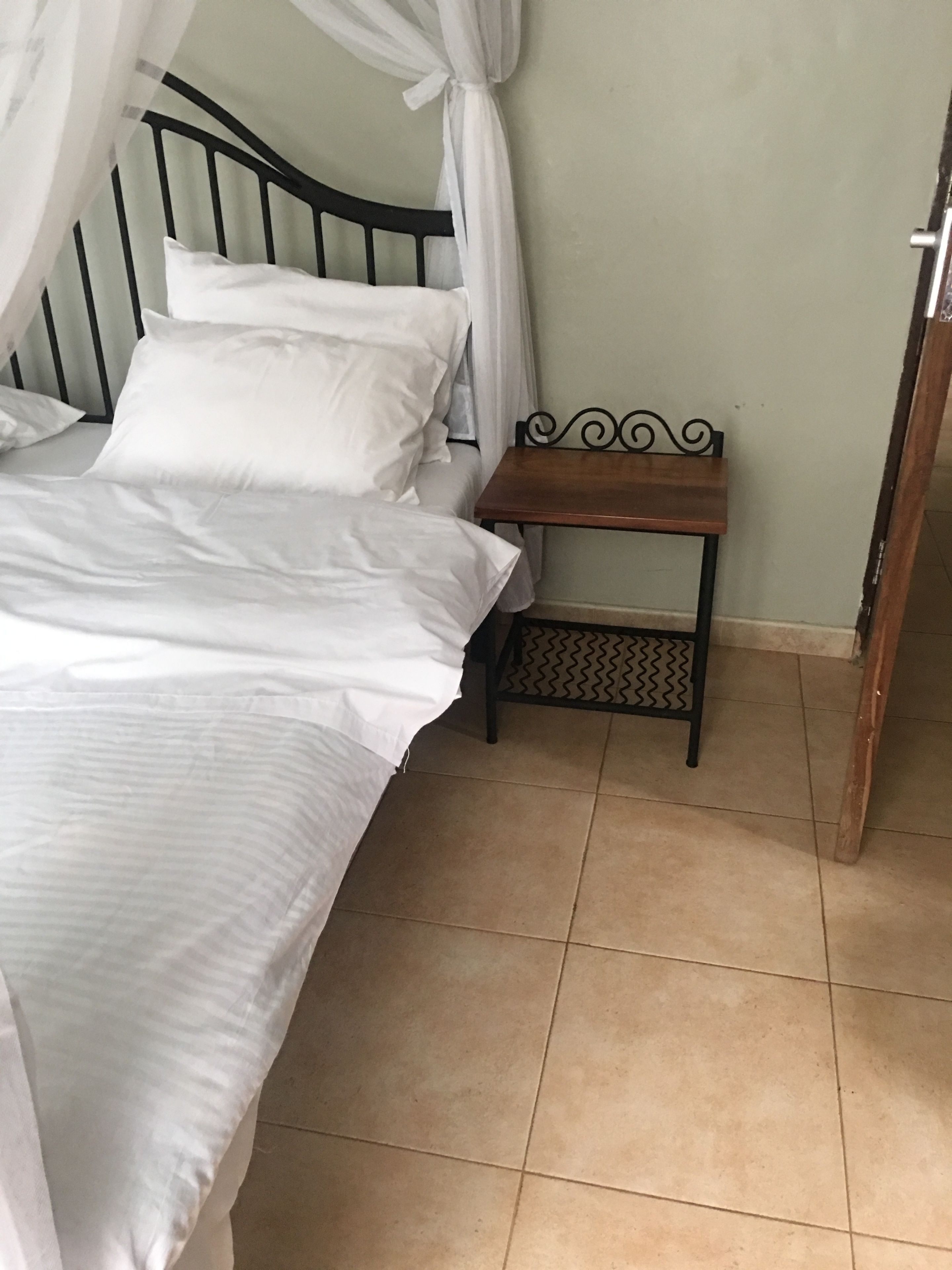 Cama com roupa de cama branca e um dossel, uma pequena mesa de cabeceira de madeira e um chão de azulejos.