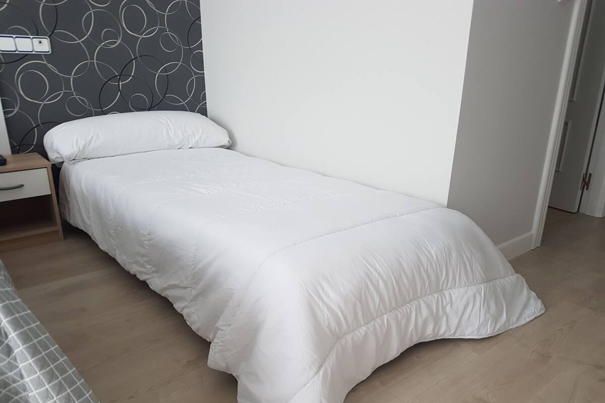 Uma cama de solteiro com edredom branco num quarto com chão de madeira e parede cinza estampada.
