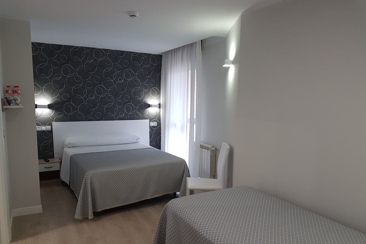 Quarto de hotel com duas camas, um radiador, uma cadeira branca e uma parede com padrão circular.