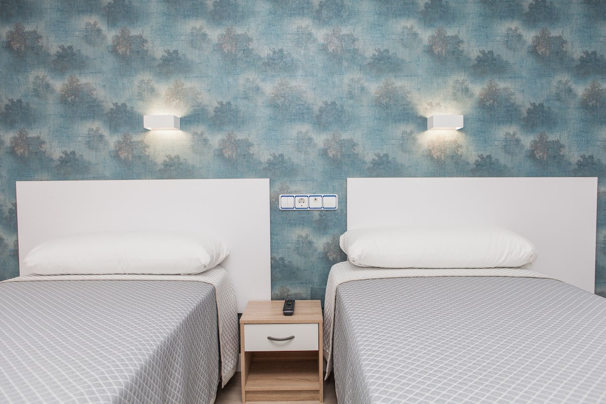 Quarto de hotel com duas camas de solteiro, uma mesinha de cabeceira de madeira com gaveta e luminária fixada na parede.