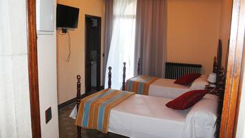 Quarto de hotel com duas camas, uma televisão fixada na parede, um radiador e uma janela com cortinas.