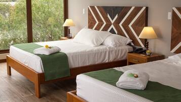 Quarto de hotel com duas camas, cada uma com uma toalha verde e um travesseiro branco, uma cabeceira de madeira, uma mesa de cabeceira com abajur e vista para árvores através de grandes janelas.