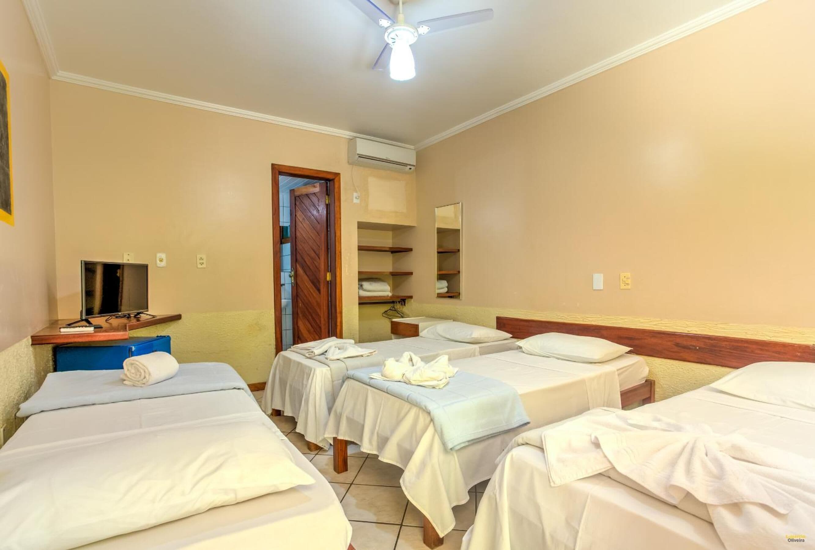 Quarto de hotel com três camas de solteiro, ventoinha no teto, televisão e armário.