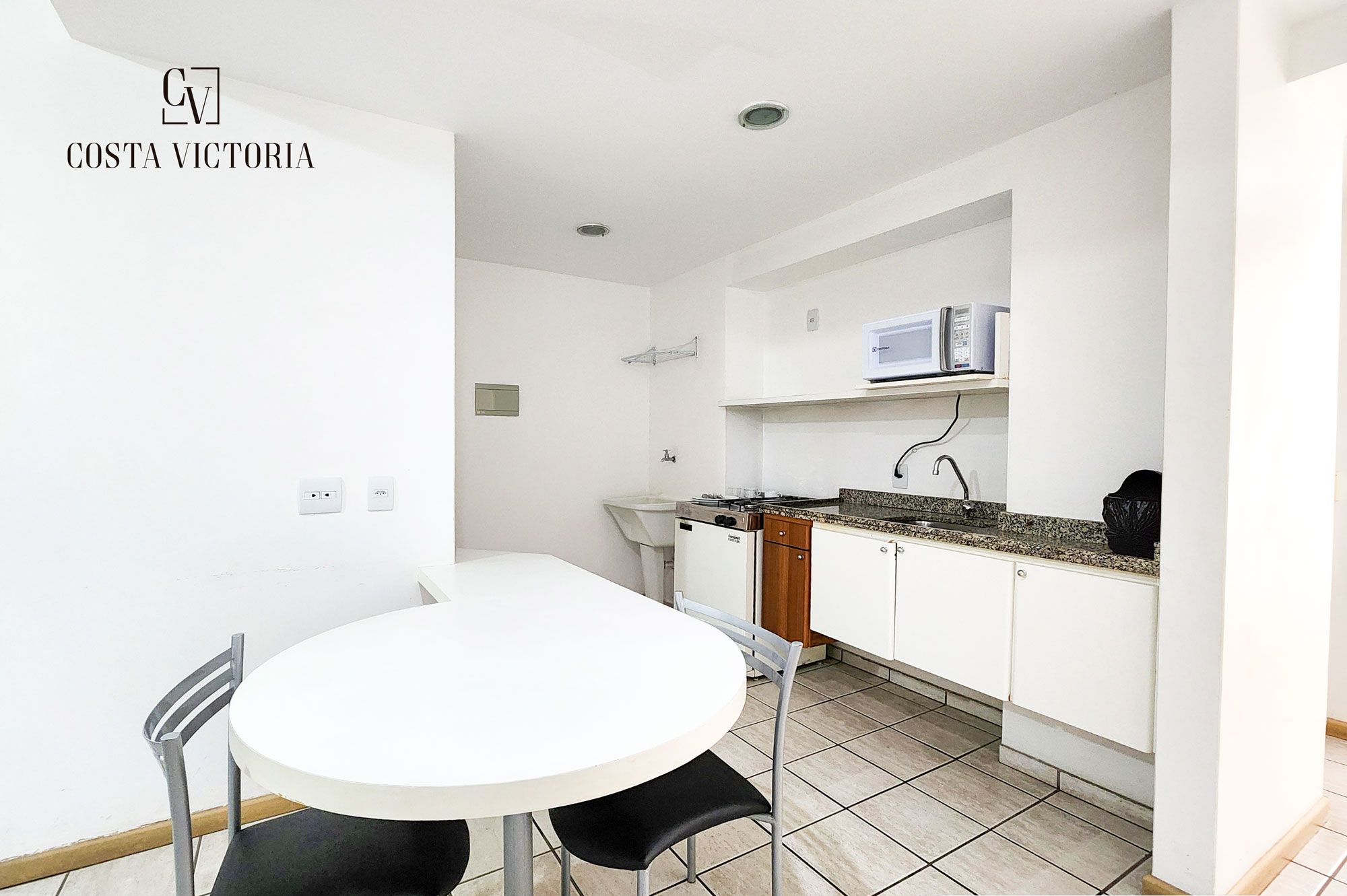 Cozinha compacta com armários brancos, micro-ondas e pia. Há uma mesa redonda com duas cadeiras.