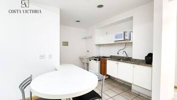 Cozinha compacta com armários brancos, micro-ondas e pia. Há uma mesa redonda com duas cadeiras.