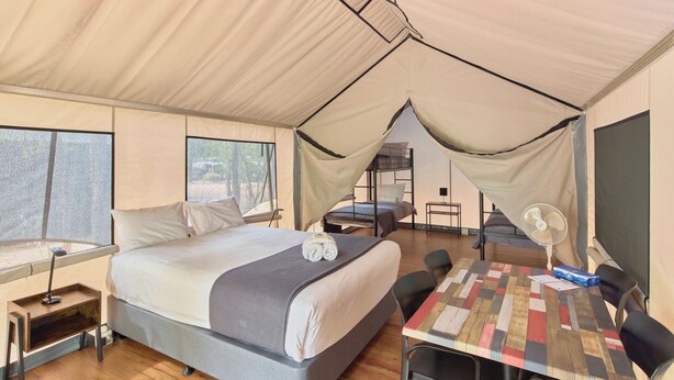 Quarto em acomodação tipo tenda, com uma cama, uma mesa com cadeiras, um ventilador e uma janela.