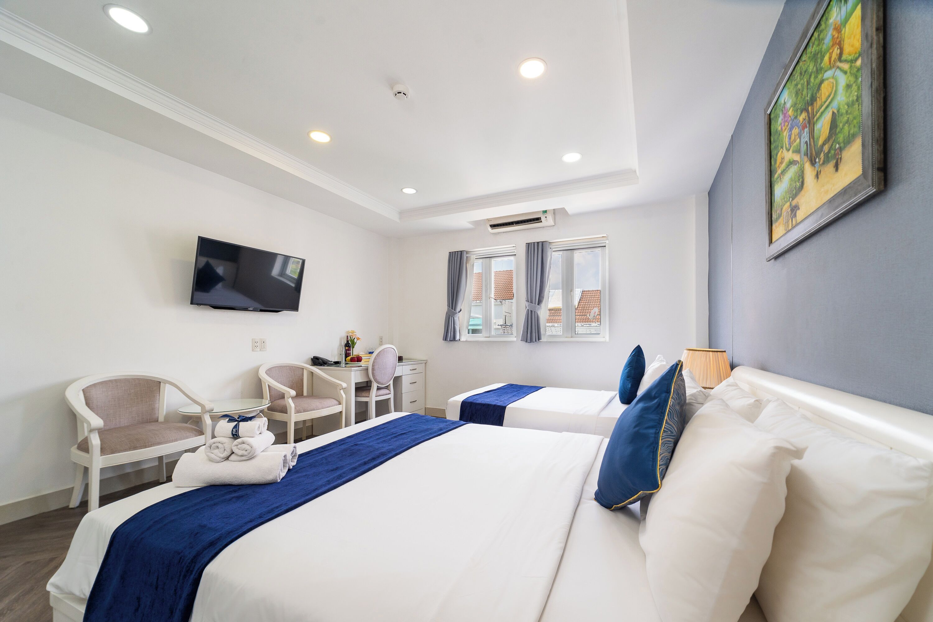 Quarto de hotel com duas camas, uma escrivaninha, cadeiras, televisão e um quadro na parede.
