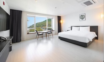 Quarto de hotel com uma cama grande, uma mesa com telefone, uma cadeira, uma mesinha e uma janela com vista para uma paisagem verde.