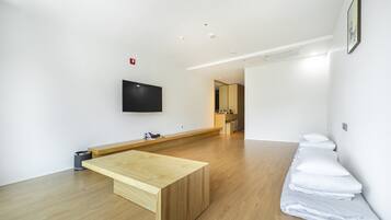 Quarto de hotel moderno com piso de madeira, TV de tela plana na parede, um banco de madeira e uma cama bem arrumada com lençóis brancos.