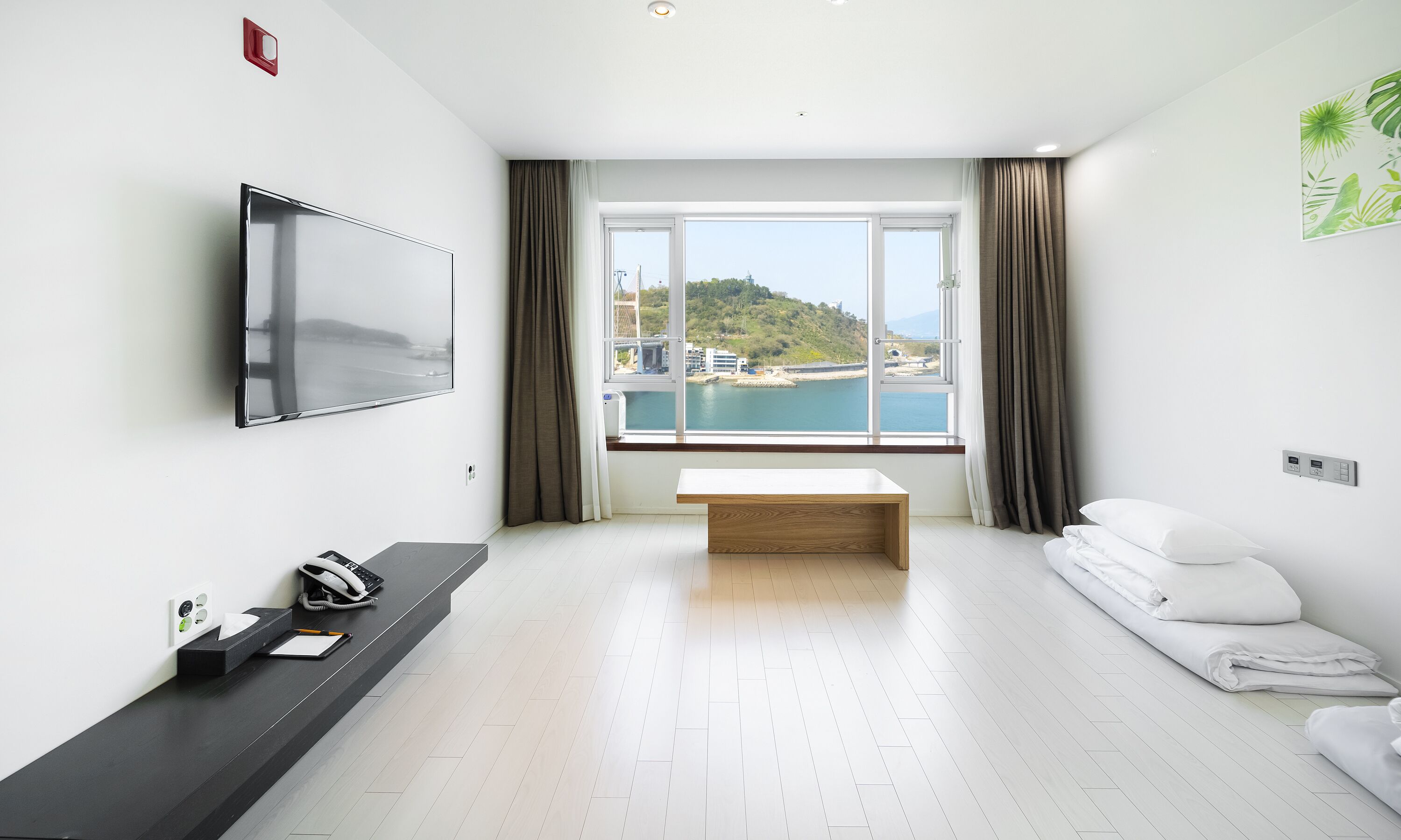 Quarto de hotel com uma janela grande, uma cama, uma mesa de cabeceira com telefone e uma televisão de tela plana na parede.
