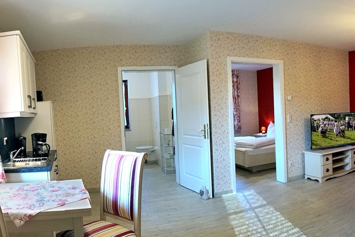 Uma sala de estar com televisão, uma parede de destaque vermelha, um candelabro e um espaço de jantar com mesa e cadeiras.