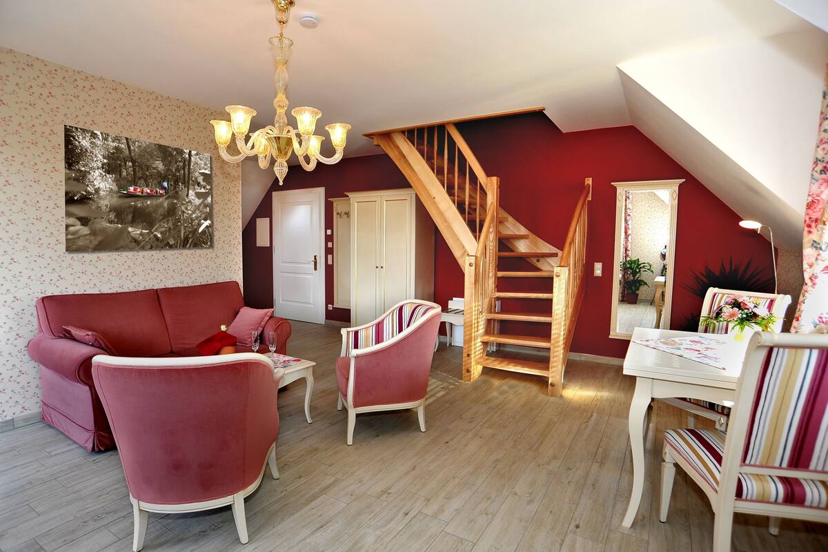 Uma sala de estar com um sofá vermelho, duas poltronas, uma escada de madeira e uma mesa de jantar com cadeiras.