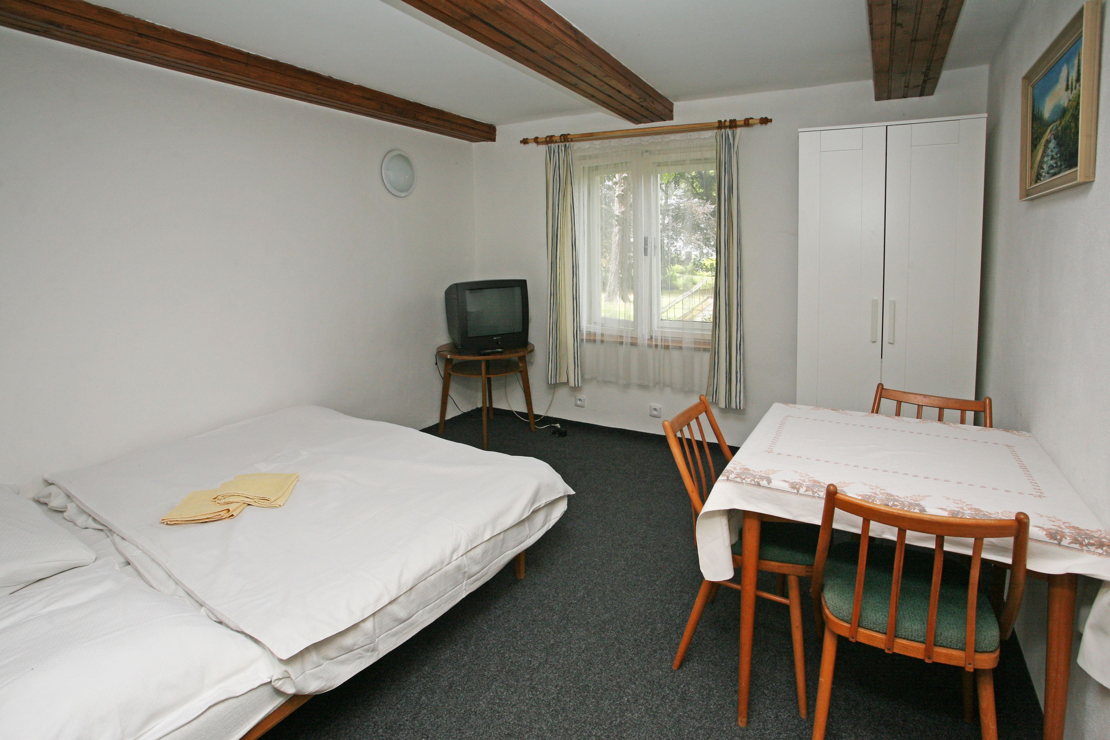 Quarto com cama, mesa e cadeiras, televisão sobre uma mesinha e janela com cortinas.