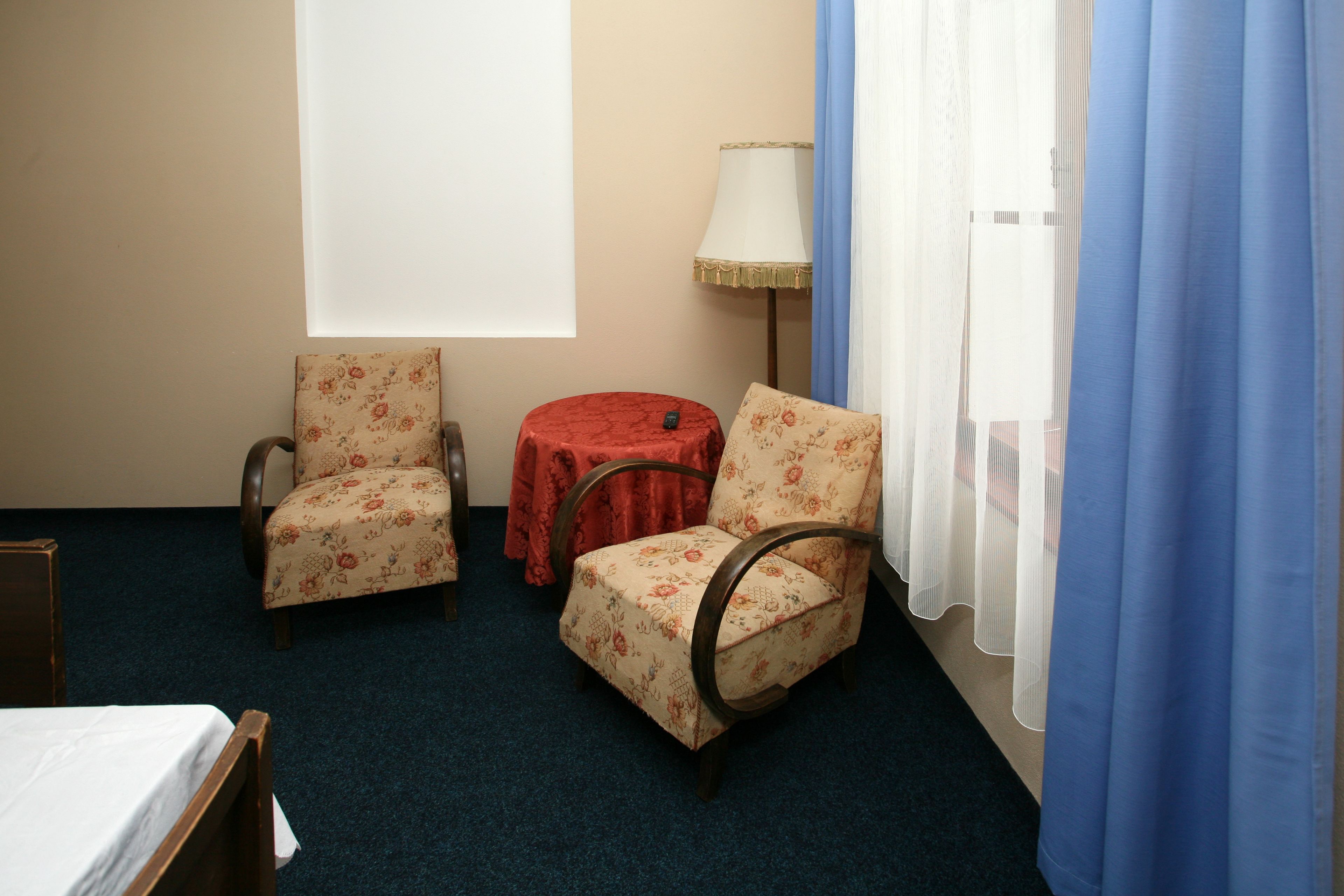 Quarto de hotel com duas poltronas estampadas com flores, uma mesa pequena vermelha, uma cama e uma janela com cortinas azuis.