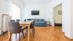 Uma sala de estar moderna com uma mesa de jantar de madeira e cadeiras, um sofá azul estampado e uma televisão de tela plana fixada na parede.