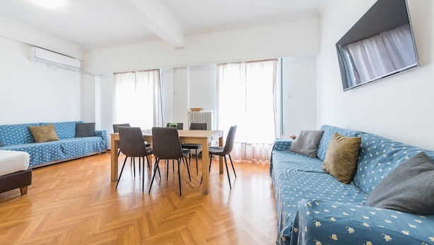 Uma sala de estar moderna com piso de madeira em padrão herringbone, uma área de jantar com mesa e cadeiras, e uma grande janela com cortinas leves.