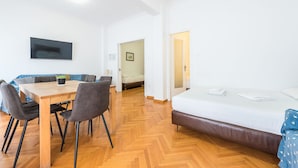 Quarto moderno e bem iluminado com mesa de jantar de madeira, cadeiras cinzentas, sofá e cama.