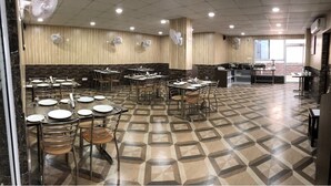 Uma ampla sala de jantar com várias mesas e cadeiras, um chão estampado e uma área de cozinha ao fundo.