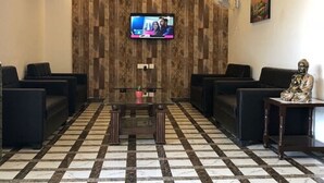 Uma área de recepção com piso xadrez, dois sofás pretos, uma mesa de centro de vidro, uma parede de destaque em madeira, uma TV montada e uma estátua em uma mesinha lateral.