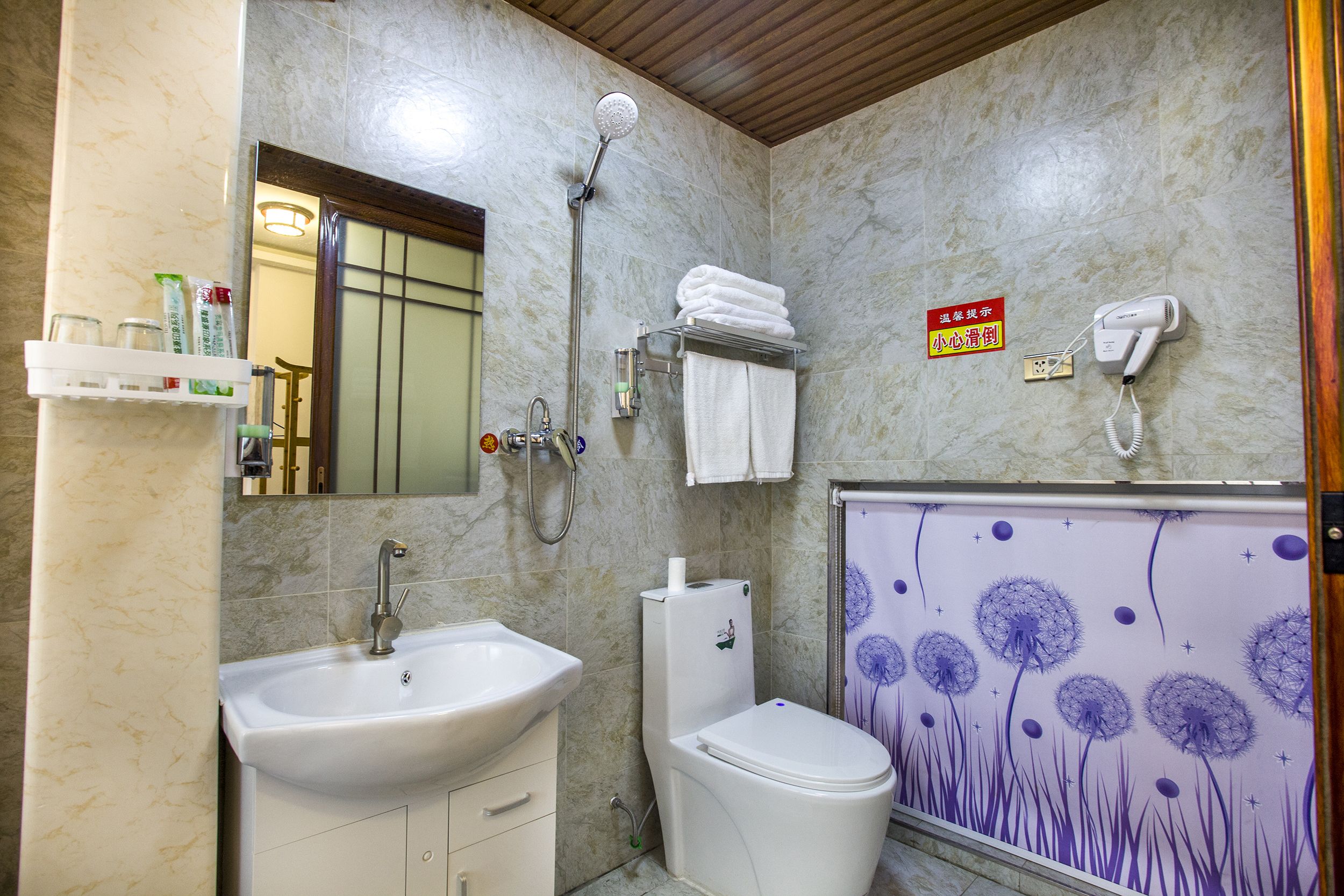 Casa de banho com lavatório, sanita e chuveiro. Há um porta-toalhas e um sinal a indicar um telefone.