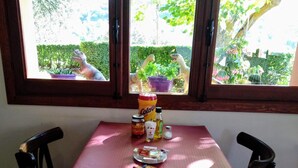 Mesa de jantar posta com pratos, copos e alimentos, com vista para um jardim e estátuas pela janela.