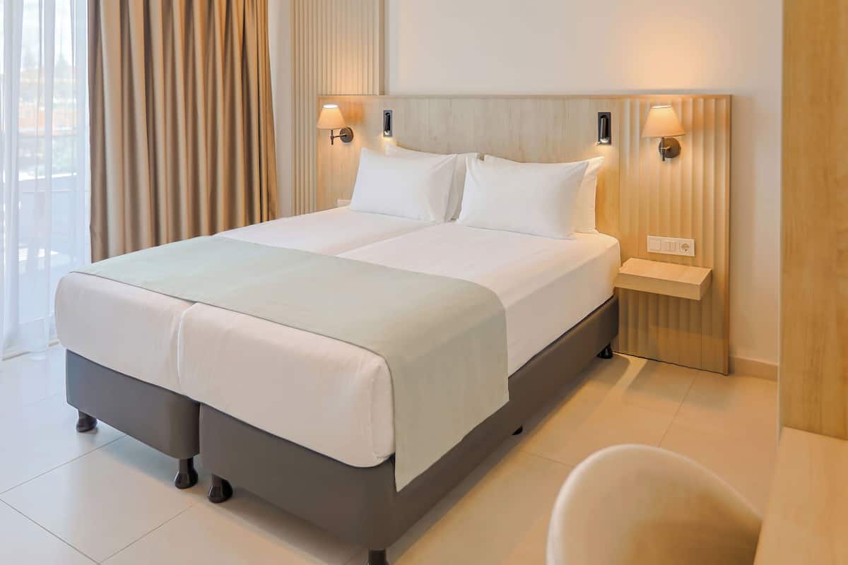 Uma cama de casal com colcha clara, duas luminárias de cabeceira, um criado-mudo com abajur e uma cadeira.
