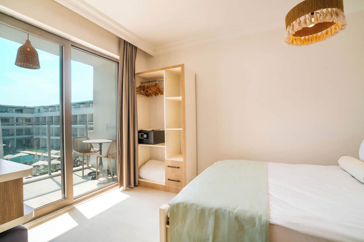 Quarto de hotel com uma grande porta de vidro deslizante que dá para uma varanda com piscina, cama com manta verde, guarda-roupa de madeira e luminária pendente.