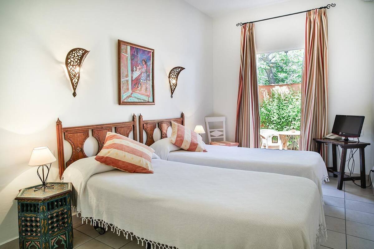 Quarto de hotel com duas camas, uma escrivaninha com computador, uma mesinha de cabeceira com abajur e um quadro na parede.