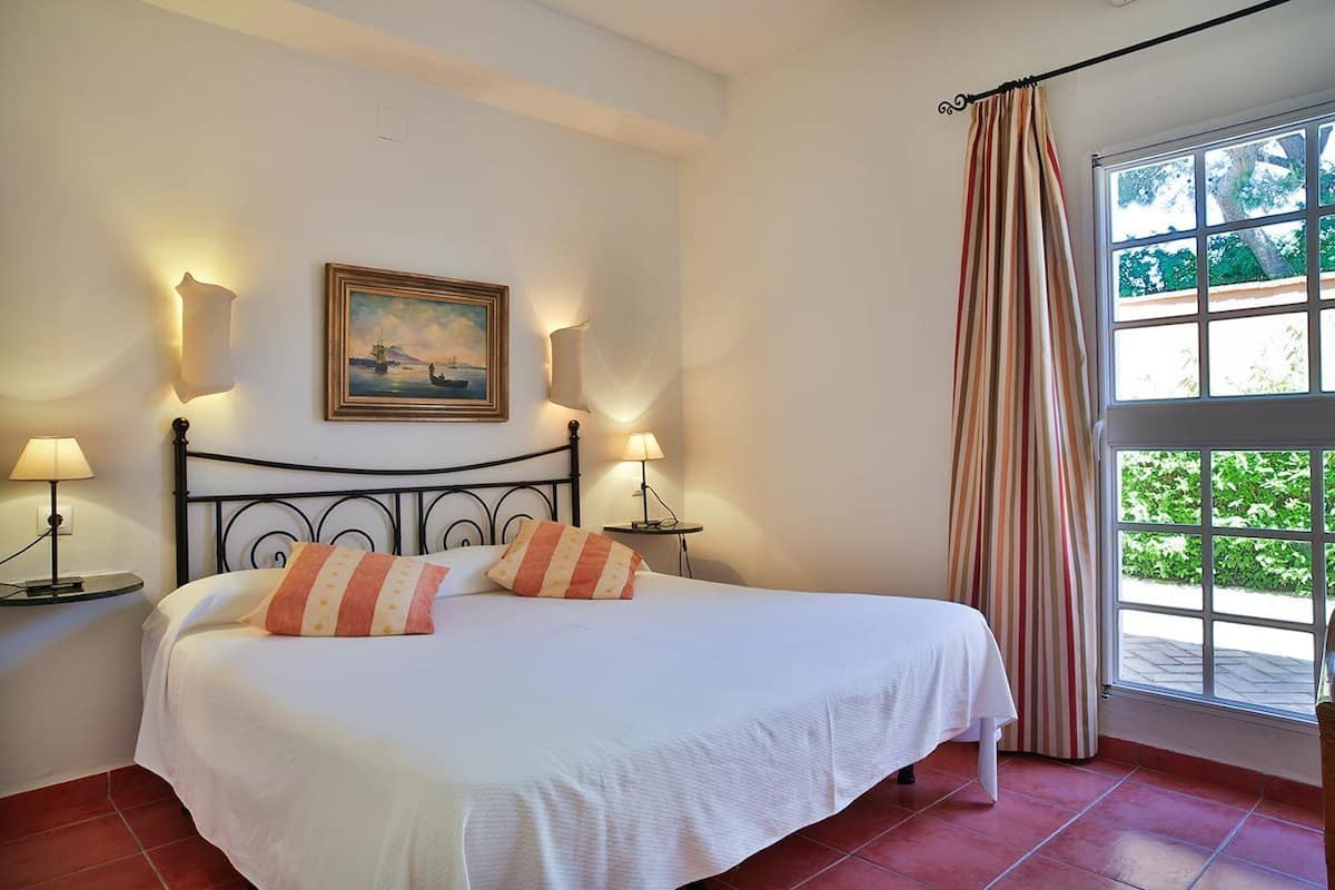 Um quarto com uma cama, duas luminárias de cabeceira, um quadro na parede e uma janela com cortinas.