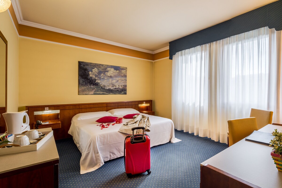 Quarto de hotel com cama, escrivaninha, cadeira, mala, abajur, televisão e um quadro na parede.