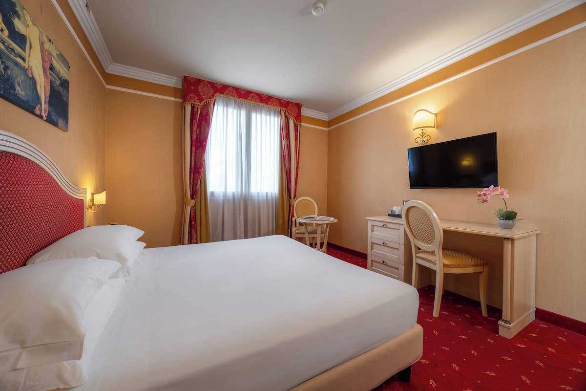 Quarto de hotel com uma cama grande, uma escrivaninha com cadeira, televisão e janela com cortinas.