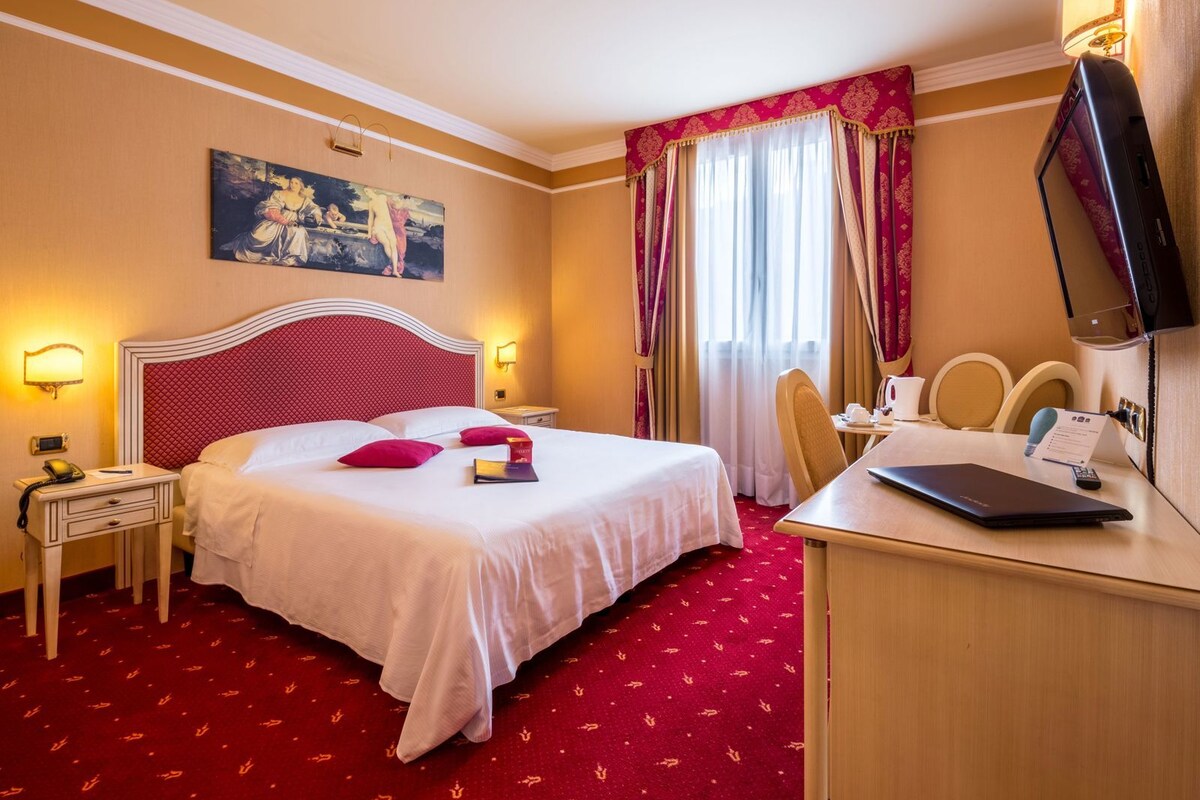 Quarto de hotel com uma cama grande, uma escrivaninha com um laptop, uma televisão e um quadro na parede.