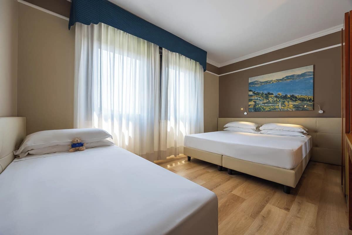 Quarto de hotel com duas camas, uma janela grande com cortinas translúcidas, um quadro na parede e piso de madeira.