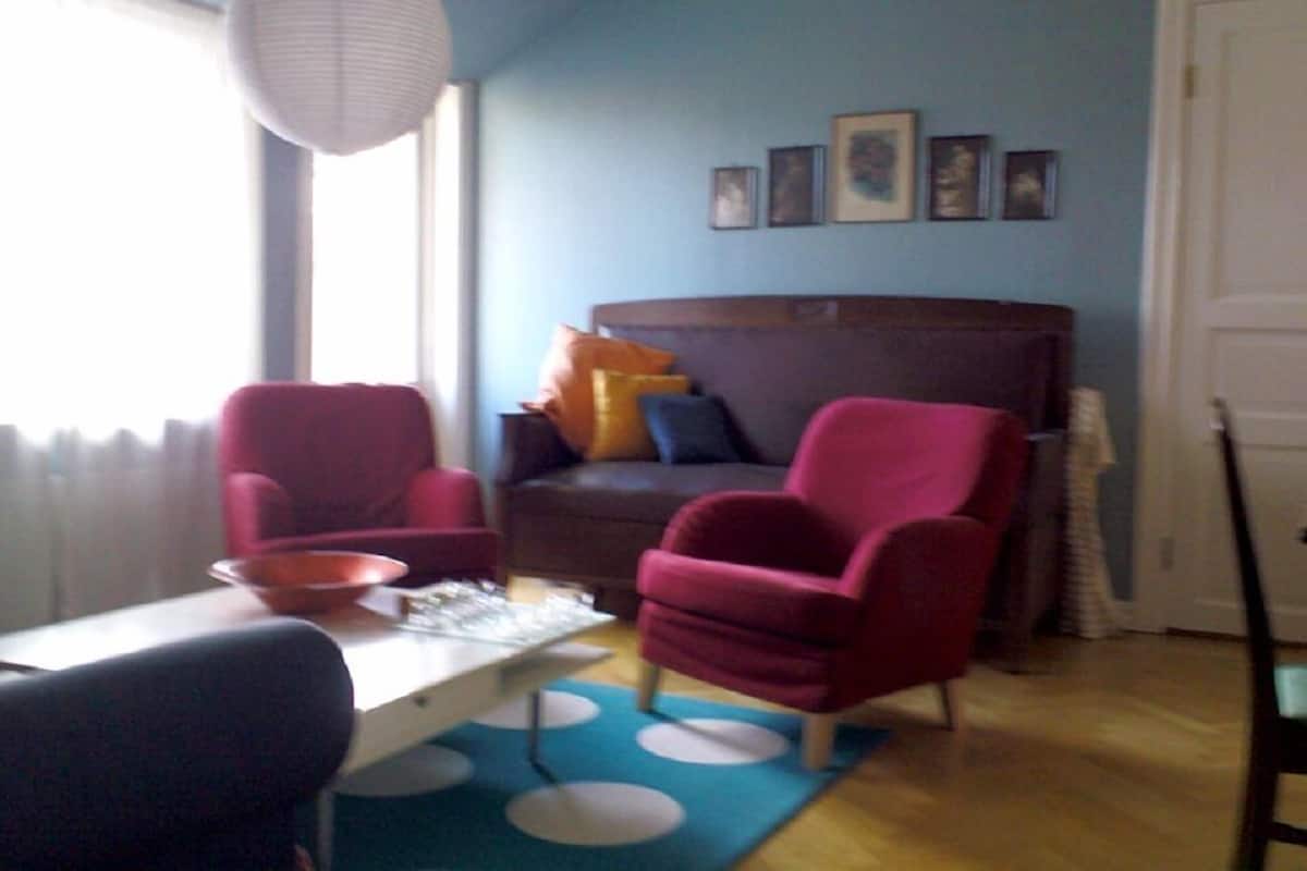 Uma sala de estar com parede azul, piso de madeira, poltrona vermelha, sofá azul com almofadas coloridas e ventilador de teto branco.