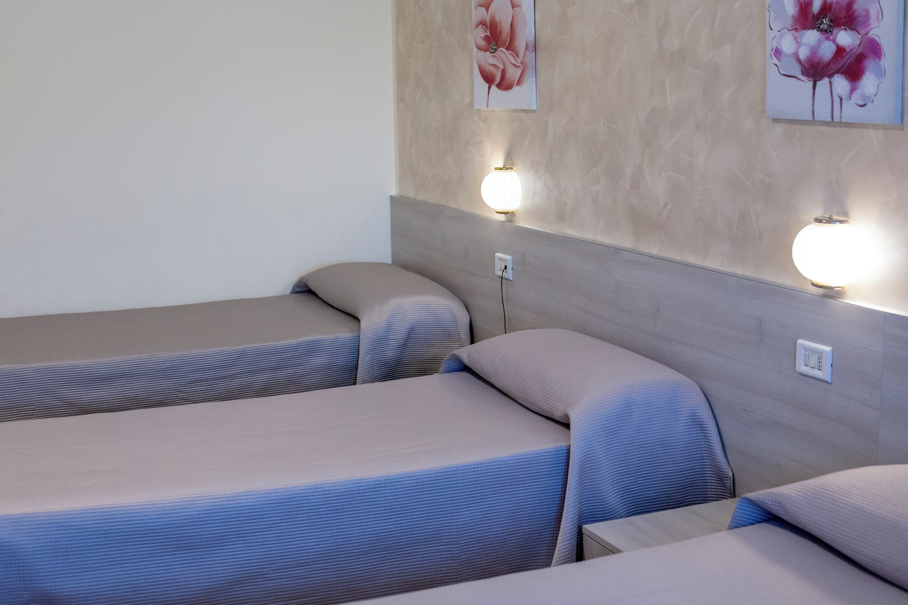 Quarto de hotel com duas camas de solteiro, cada uma com travesseiro e cabeceira. Há luminárias fixadas na parede em ambos os lados das camas.