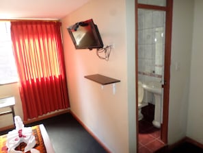 Quarto de hotel com televisão na parede, um pequeno armário, janela com cortinas vermelhas e vista para o banheiro.