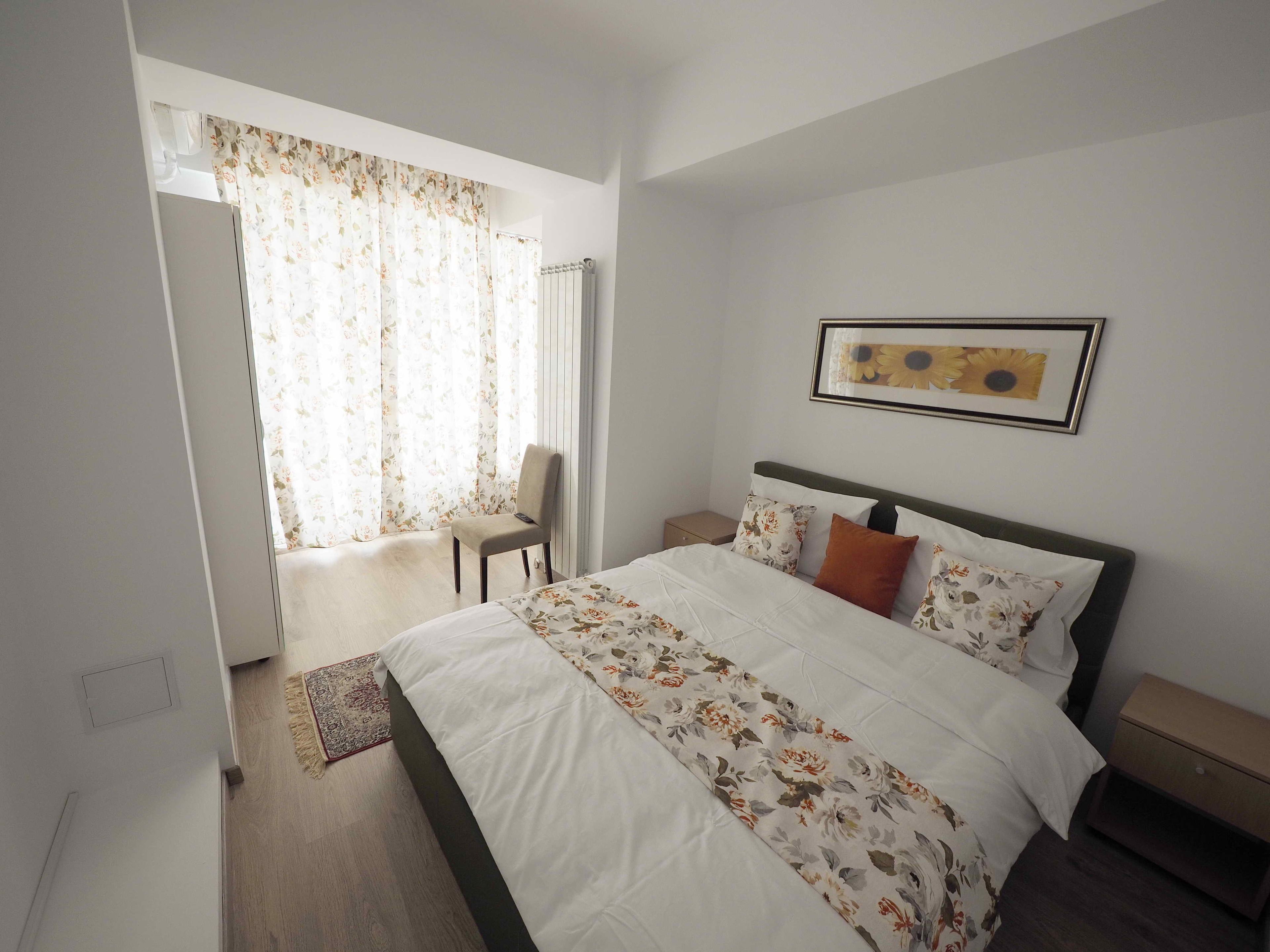 Quarto com cama, cadeira, mesa de cabeceira, radiador e janela com cortinas.