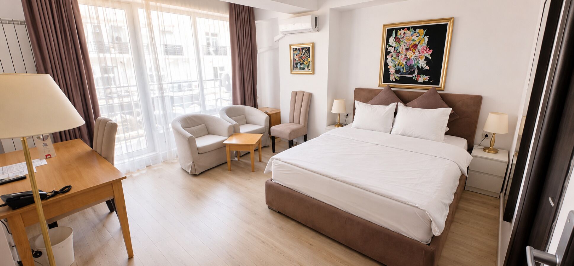Quarto de hotel com cama, duas poltronas, escrivaninha com luminária, mesa de cabeceira com luminária e janela com cortinas.
