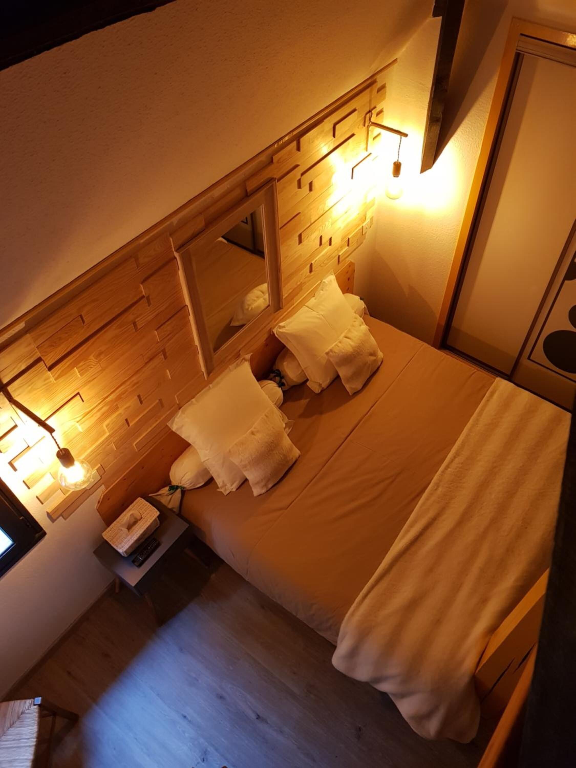 Quarto com cabeceira de madeira, cama com roupa de cama branca, mesa de cabeceira com abajur e espelho.