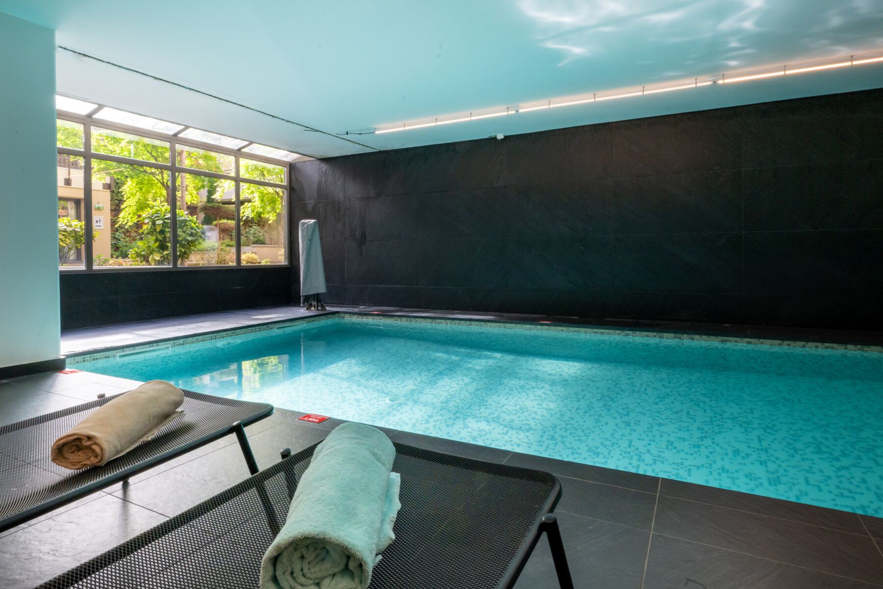 Piscina interna com água azul cristalina, parede revestida de azulejos escuros e janela com vista para o verde lá fora.