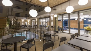 Amplo espaço interno com várias mesas e cadeiras, uma piscina e teto alto com luminárias pendentes.