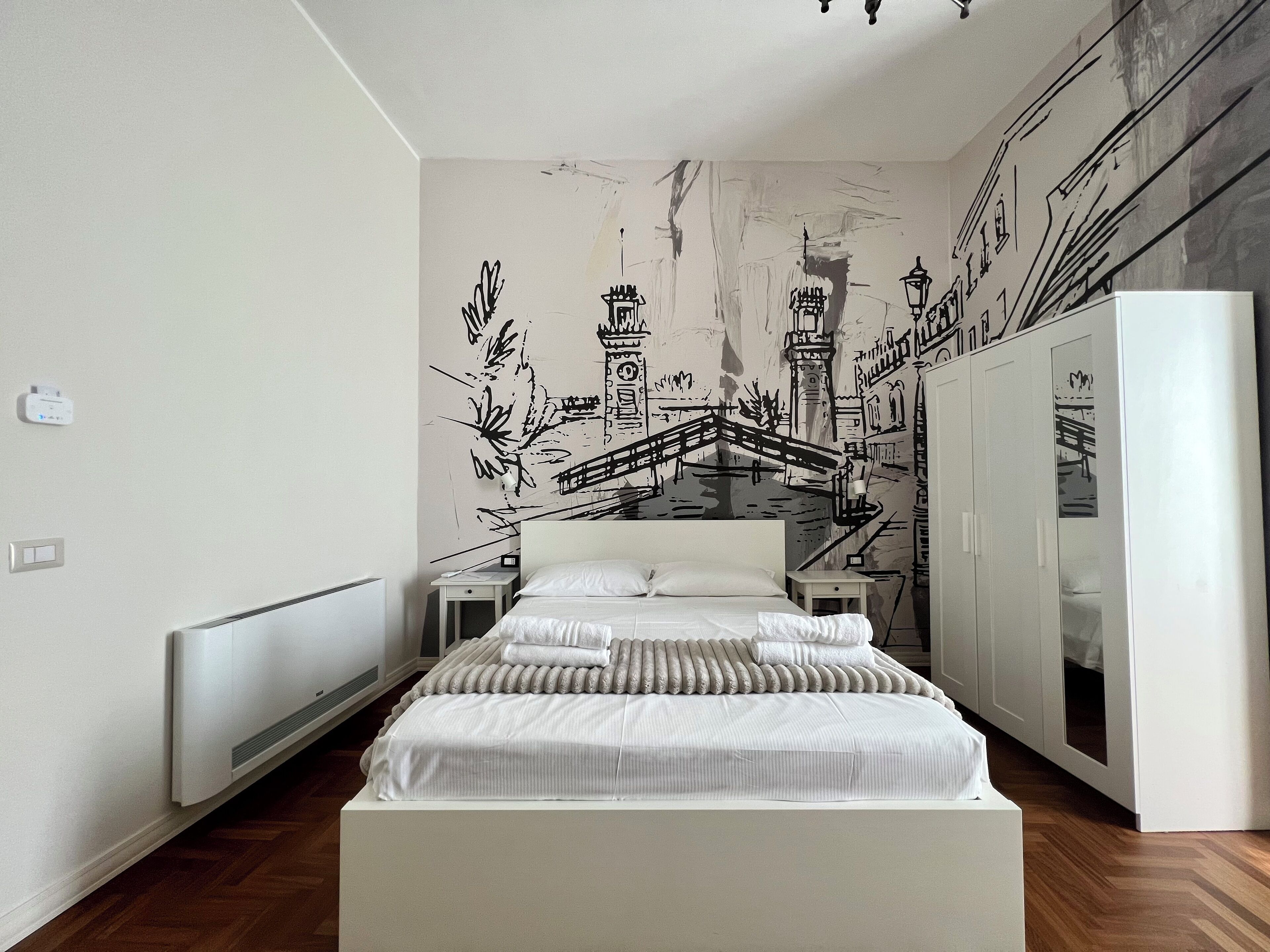 Quarto moderno com cama, mesas de cabeceira, radiador e um mural na parede com vista para a cidade.