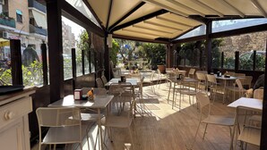 Um café ao ar livre com mesas e cadeiras, grandes janelas e vista para a rua.