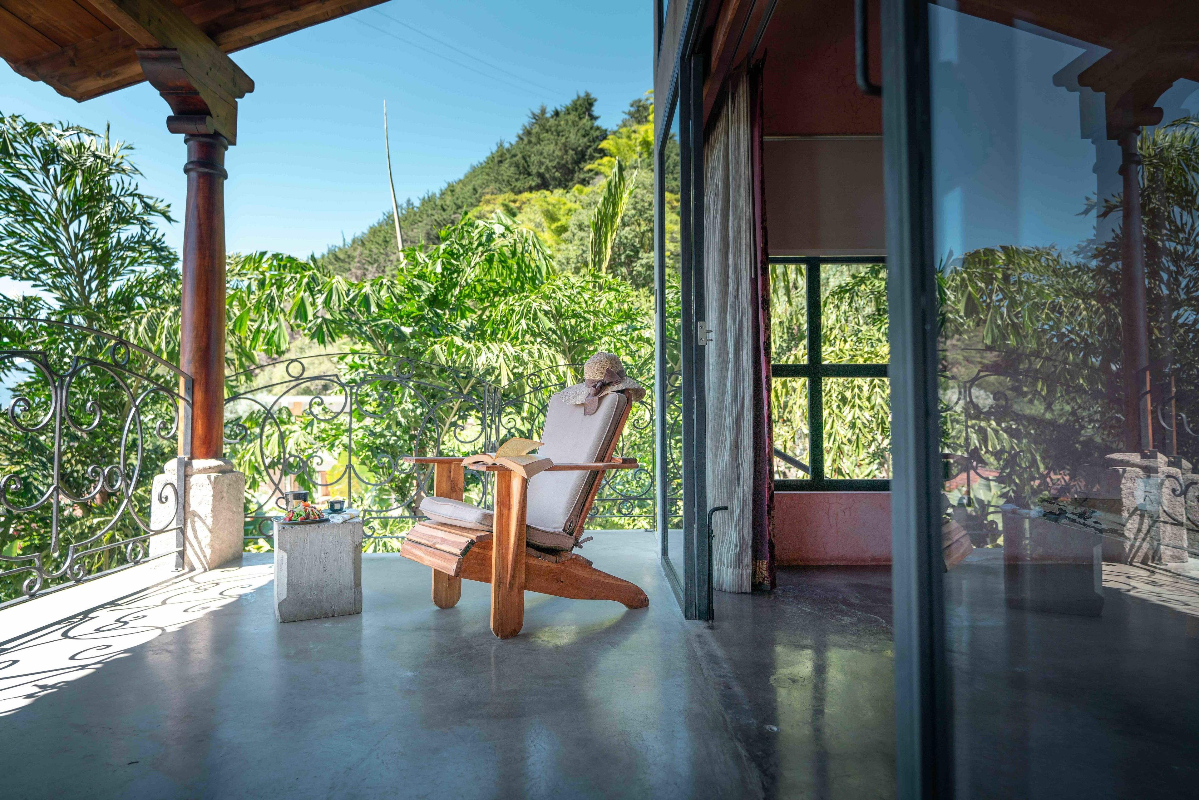 Uma cadeira de madeira em varanda, com base de concreto, com vista para um jardim exuberante e uma montanha ao fundo.
