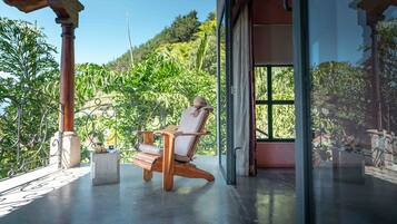 Uma cadeira de madeira em varanda, com base de concreto, com vista para um jardim exuberante e uma montanha ao fundo.