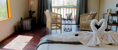 Quarto de hotel com uma cama, uma mesa pequena com uma garrafa, duas cadeiras de vime, uma varanda com vista e um vaso de plantas.