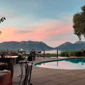 Uma área à beira da piscina com cadeiras e mesas, com vista para um lago e montanhas.