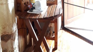 Interior de madeira com uma mesa onde se encontram uma cafeteira, xícaras e uma pequena caixa. Uma televisão está sobre a mesa, e uma janela revela vista para o exterior.