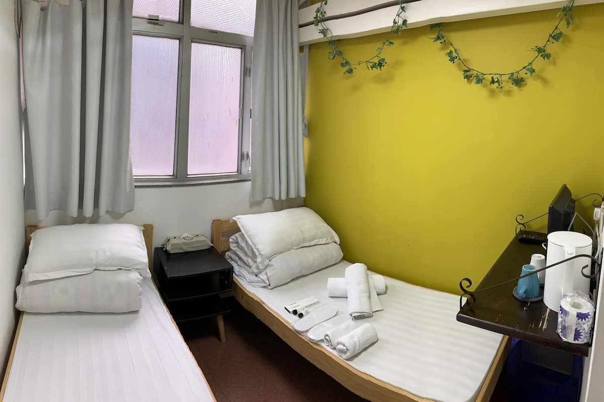Quarto individual com janela, escrivaninha com computador e pequena mesa com artigos de higiene.